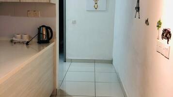 Uma cozinha moderna com armários de madeira, uma chaleira e uma cafeteira na bancada. Um corredor com piso ladrilhado leva a um quarto com uma porta.