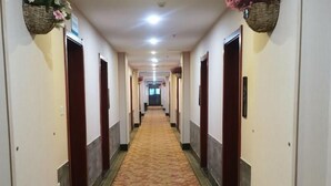 Corredor de hotel com piso coberto por carpete, portas em ambos os lados e cestos suspensos com flores.