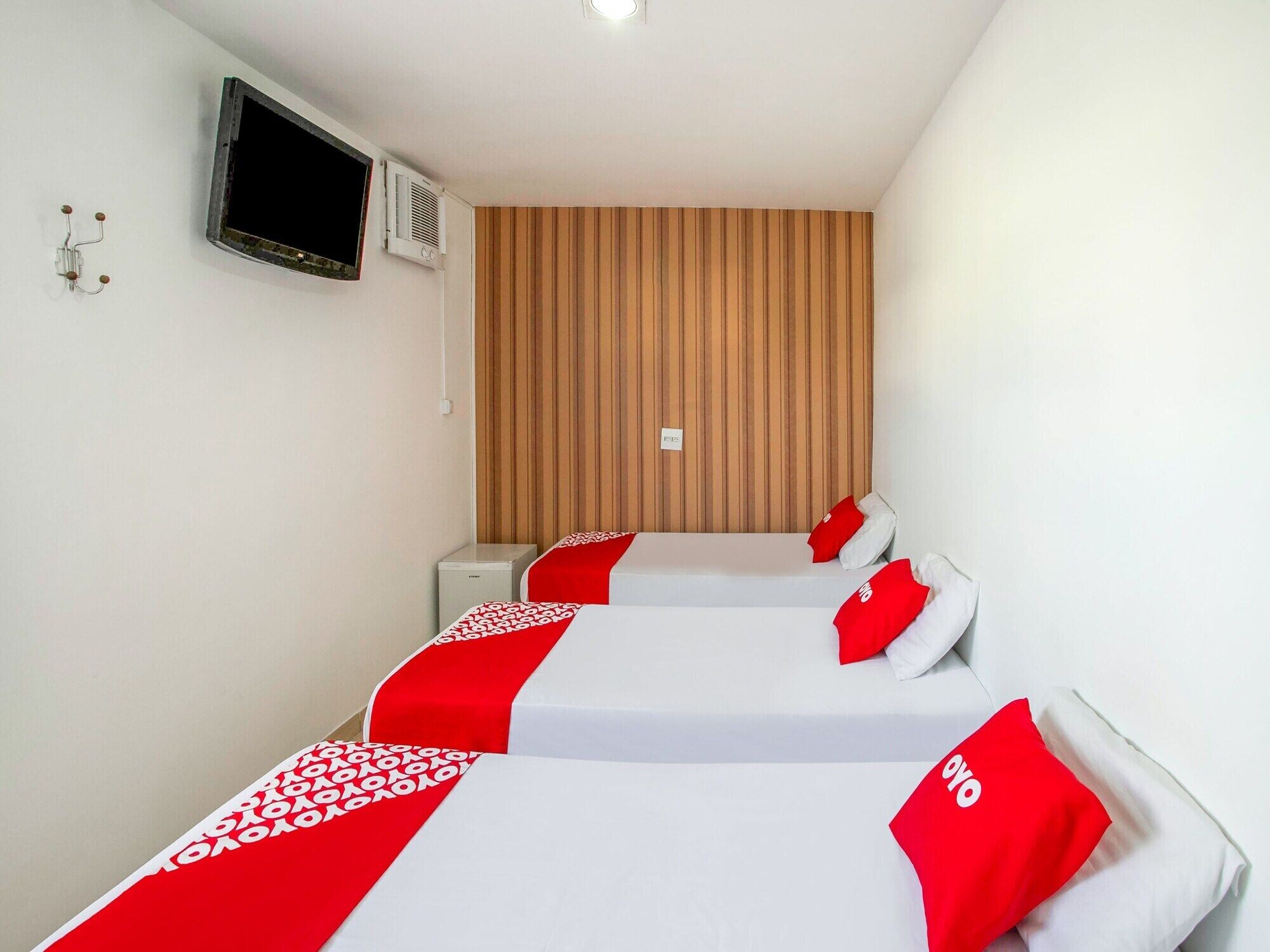 Quarto de hotel com duas camas, cada uma com roupa de cama vermelha e branca, uma televisão na parede e painel de parede de madeira.