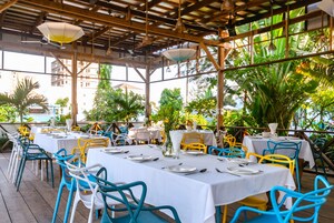 Restaurante ao ar livre com mesas e cadeiras, coberto por um telhado de madeira, rodeado por vegetação.