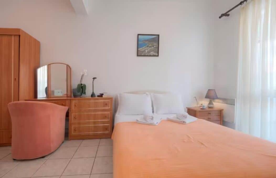 Um quarto com cama, penteadeira de madeira, cadeira rosa, mesa de cabeceira, luminária e quadro na parede.