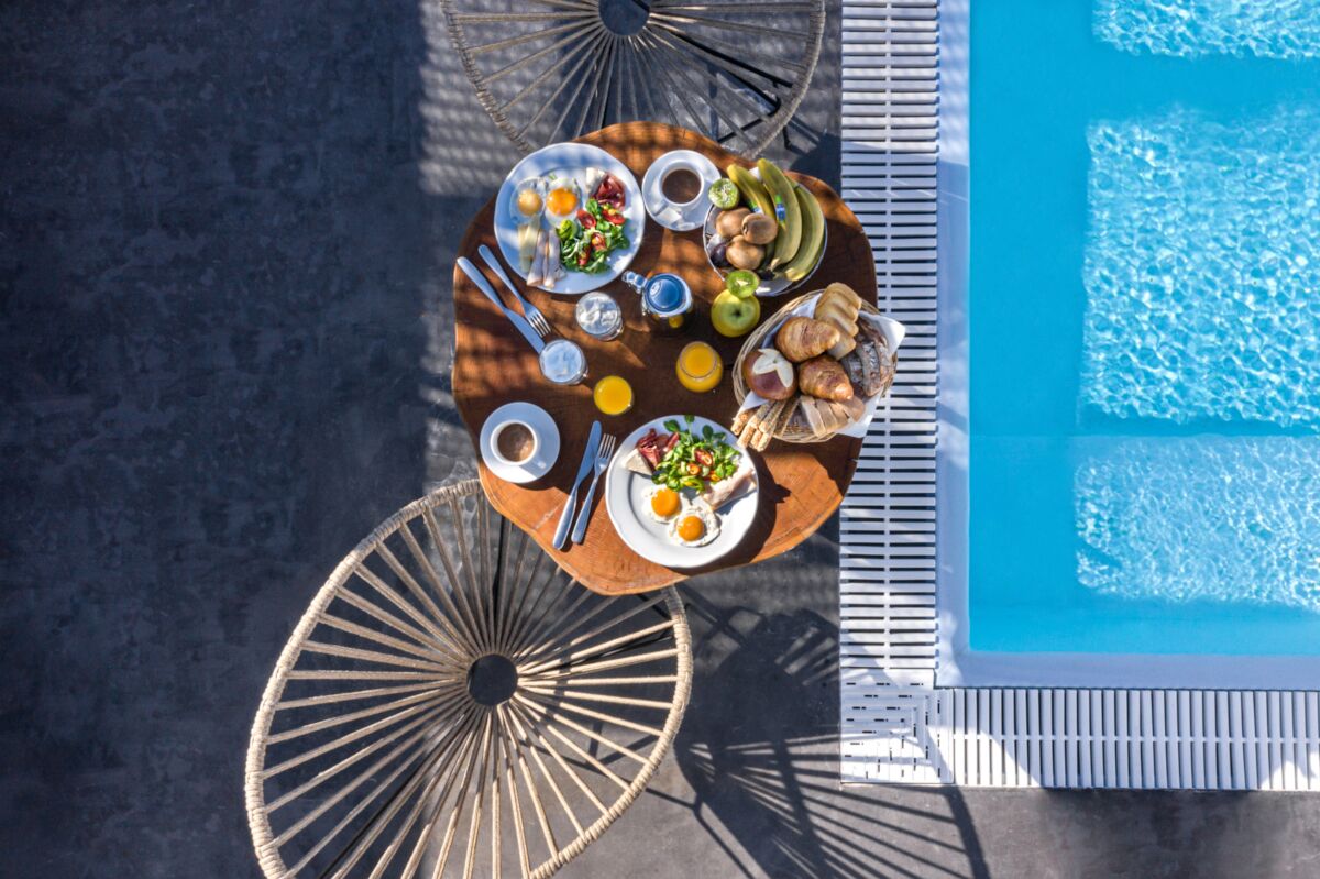 Uma área de refeições ao ar livre com vista para a piscina, apresentando uma mesa redonda com pratos de comida, uma xícara de café e uma cesta de pão.