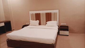 Cama bem arrumada com estrutura de madeira e roupa de cama branca, mesa de cabeceira com abajur e um pequeno conjunto de gavetas ao lado da cama.