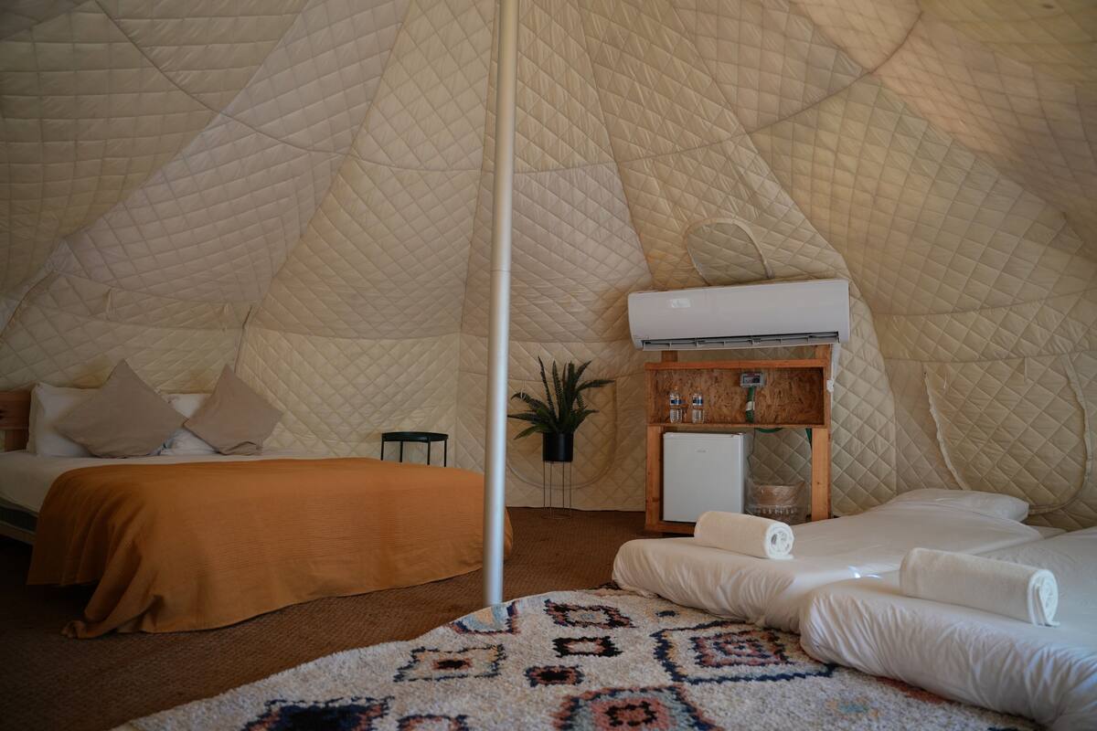 Quarto tipo tenda com uma cama, uma mesa pequena com uma planta e um aparelho de ar condicionado.