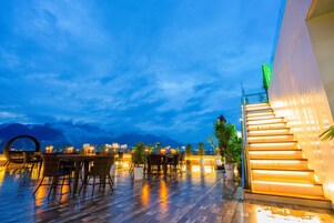 Uma área de refeições ao ar livre com mesas e cadeiras, uma escadaria com degraus iluminados e vista para as montanhas ao fundo.