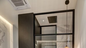 Quarto moderno com uma cama grande, box de vidro para o chuveiro, luminária pendente e uma pequena planta em vaso.