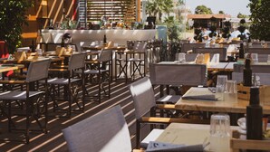 Restaurante ao ar livre com mesas e cadeiras de madeira, área de bar e vista para palmeiras e edifícios ao fundo.
