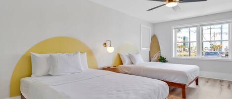 Quarto de hotel com duas camas, um ventilador de teto, uma janela com vista para o exterior e uma pequena planta no parapeito.