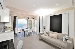 Quarto de hotel moderno com cozinha compacta, área de refeições com mesa e cadeiras, sofá e uma janela grande com vista.
