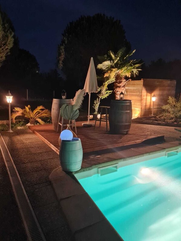 Uma área de piscina externa à noite, com um deck de madeira, um vaso de planta e uma mesa com cadeiras.