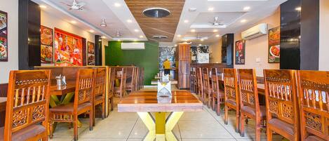 Interior de restaurante com mesas e cadeiras de madeira, uma mesa central em formato de X e ventiladores de teto.