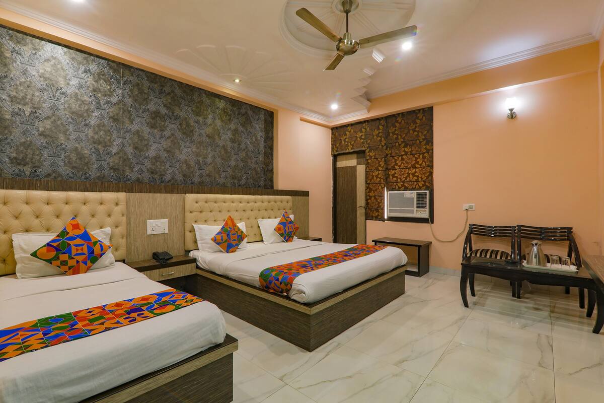 Quarto de hotel com duas camas, um ventilador de teto, um aparelho de ar condicionado, um banco de madeira e uma mesa com jarra e copos.