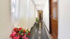 Um corredor com uma porta de madeira à direita, paredes brancas e um vaso com flores no final.