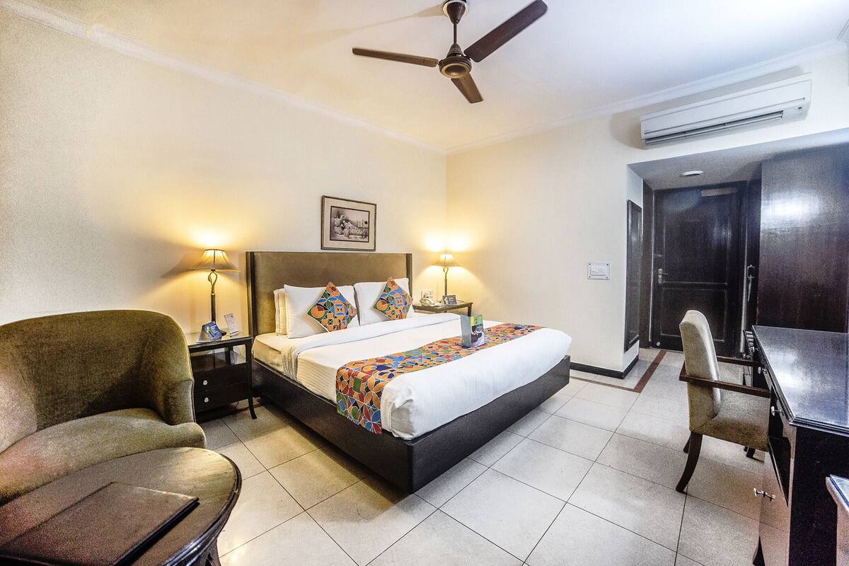 Quarto de hotel com uma cama, duas mesas de cabeceira, uma escrivaninha, uma cadeira, um ventilador de teto e um ar condicionado.