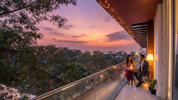 Varanda com vista para a cidade ao pôr do sol, guarda-corpo de vidro e uma mesa para duas pessoas.