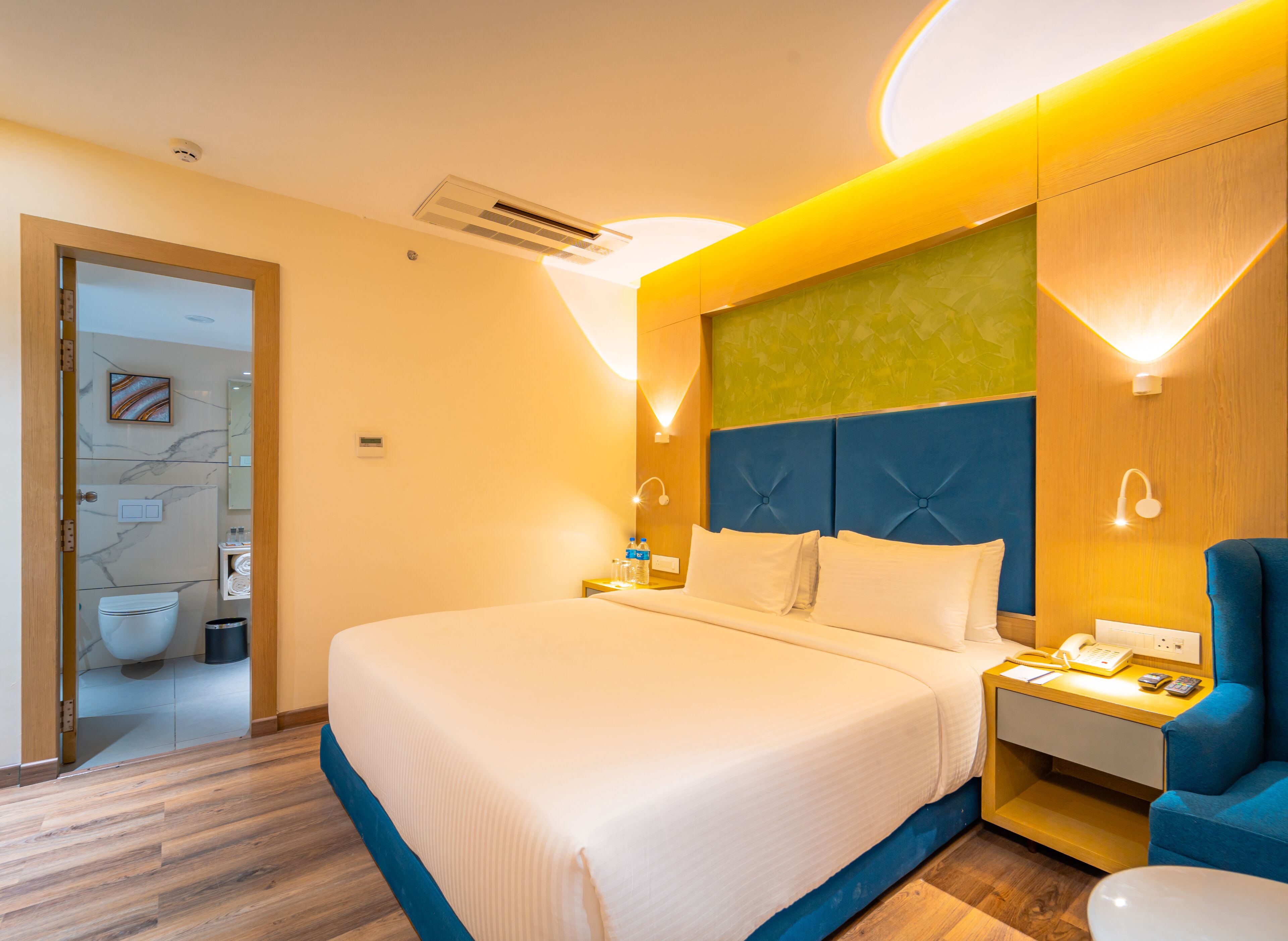 Quarto de hotel com uma cama grande, uma mesa de cabeceira com telefone e luminária, uma poltrona azul e um banheiro visível por uma porta aberta.