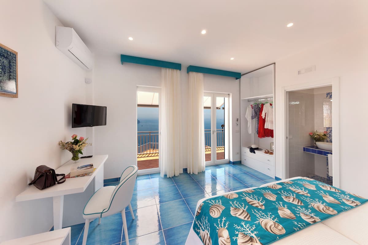 Quarto de hotel com cama, escrivaninha, cadeira, televisão, banheiro e varanda com vista.