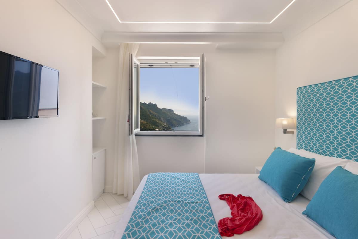 Quarto de hotel moderno com uma cama grande, televisão fixada na parede e janela com vista panorâmica da costa.