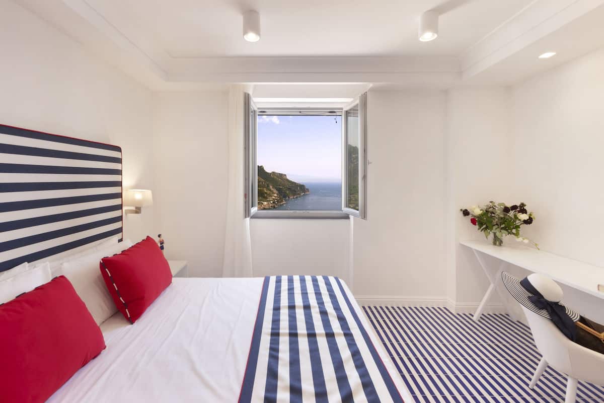 Quarto com cama, escrivaninha e janela com vista para o mar.