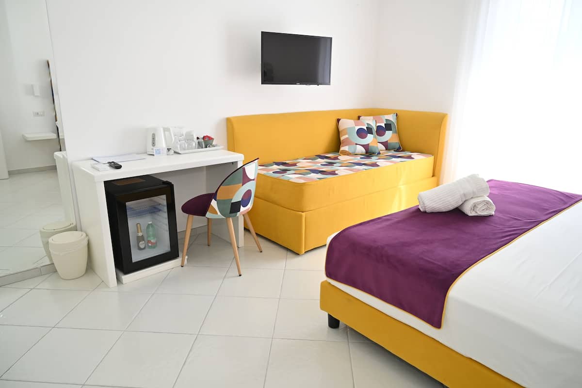 Quarto de hotel com um sofá amarelo, uma cama com manta roxa, uma escrivaninha com computador e uma TV fixada na parede.