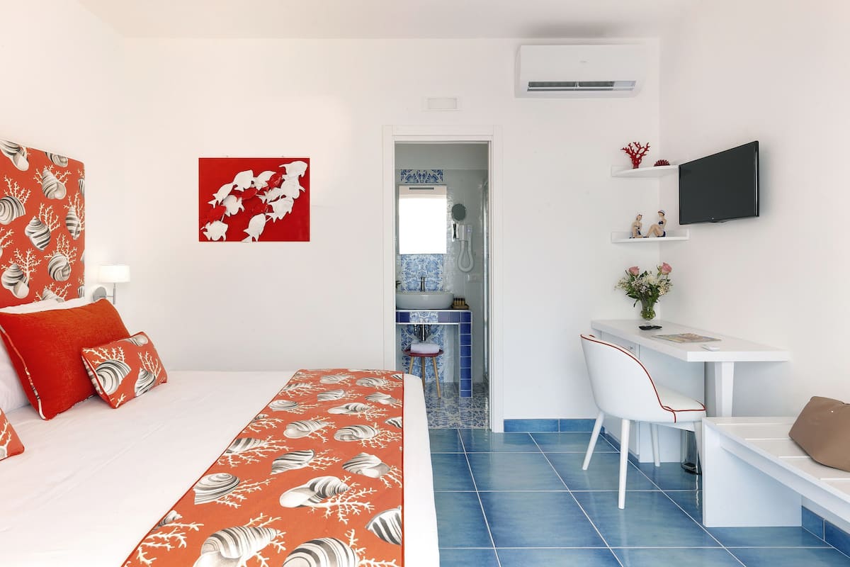 Quarto de hotel com cama, escrivaninha, cadeira, televisão e banheiro visível através de uma porta aberta.