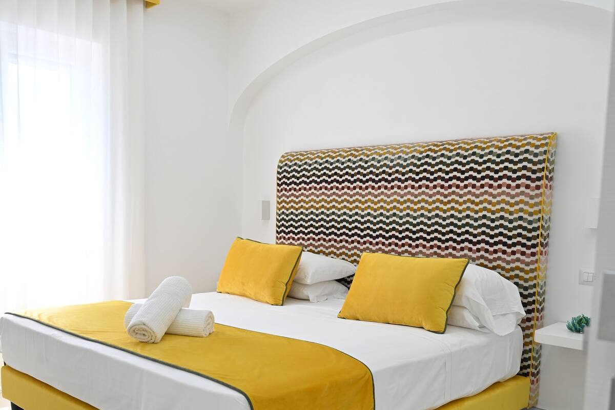 Quarto moderno com colcha amarela, roupa de cama branca e cabeceira estampada.