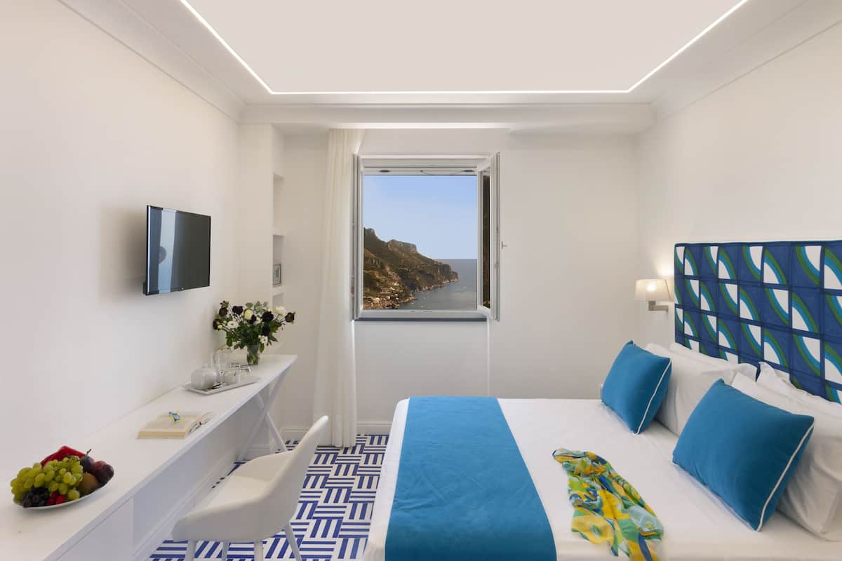 Quarto de hotel com cama, escrivaninha com uma cesta de fruta, televisão e janela com vista para a costa.