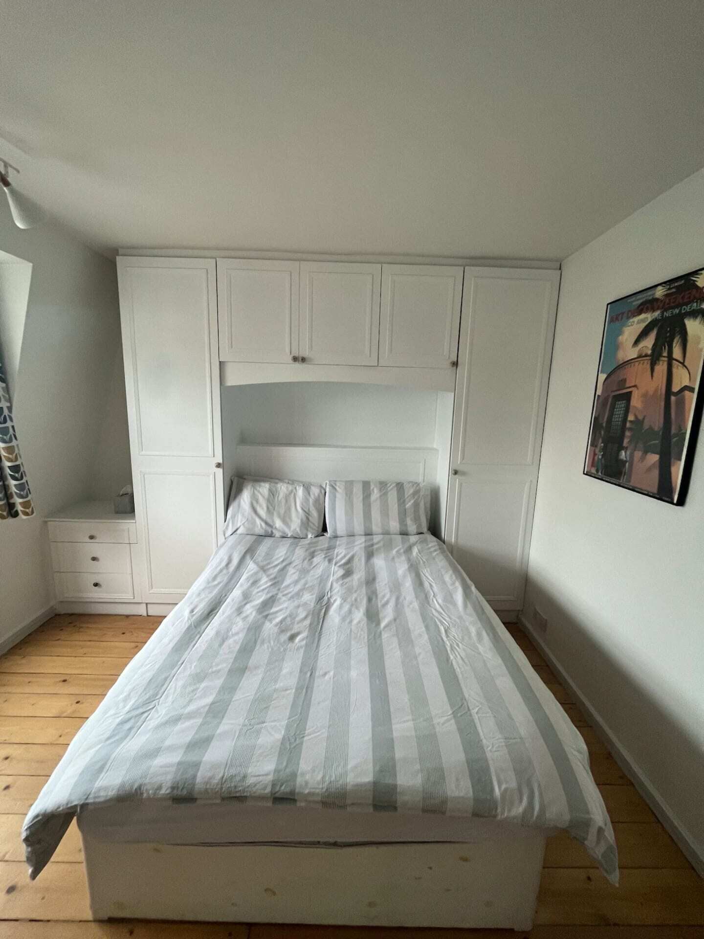 Quarto com cama, armários brancos, mesa de cabeceira e um cartaz na parede.