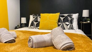 Uma cama bem arrumada com um cobertor amarelo e toalhas dobradas, um travesseiro estampado com padrão geométrico e uma mesa de cabeceira com abajur.