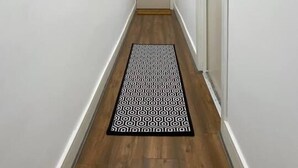 Um corredor estreito com paredes brancas, chão de madeira e tapete de entrada com padrão em preto e branco.