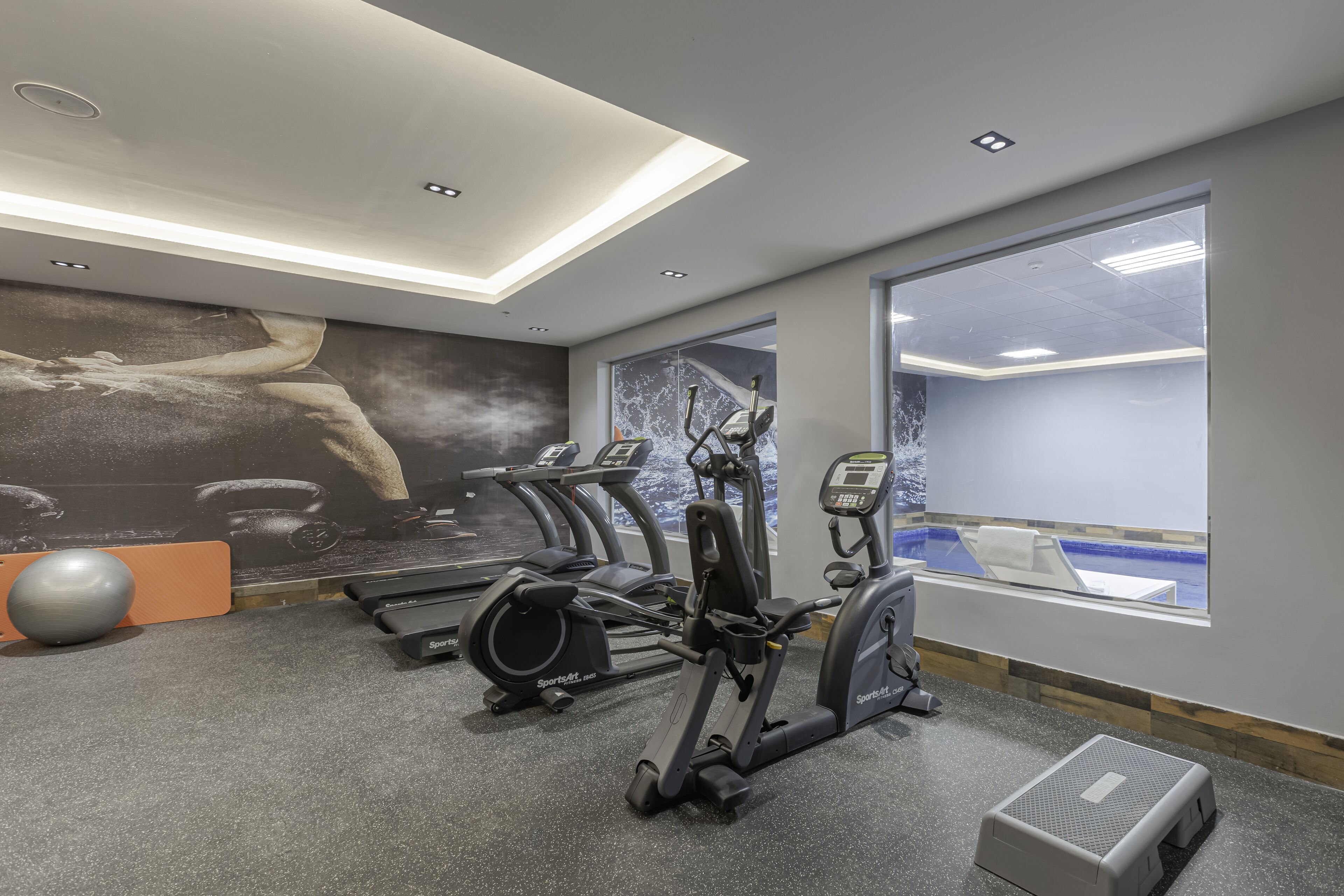 Uma academia com esteiras, elípticos e uma bicicleta ergométrica, uma janela grande com vista e um teto com iluminação embutida.