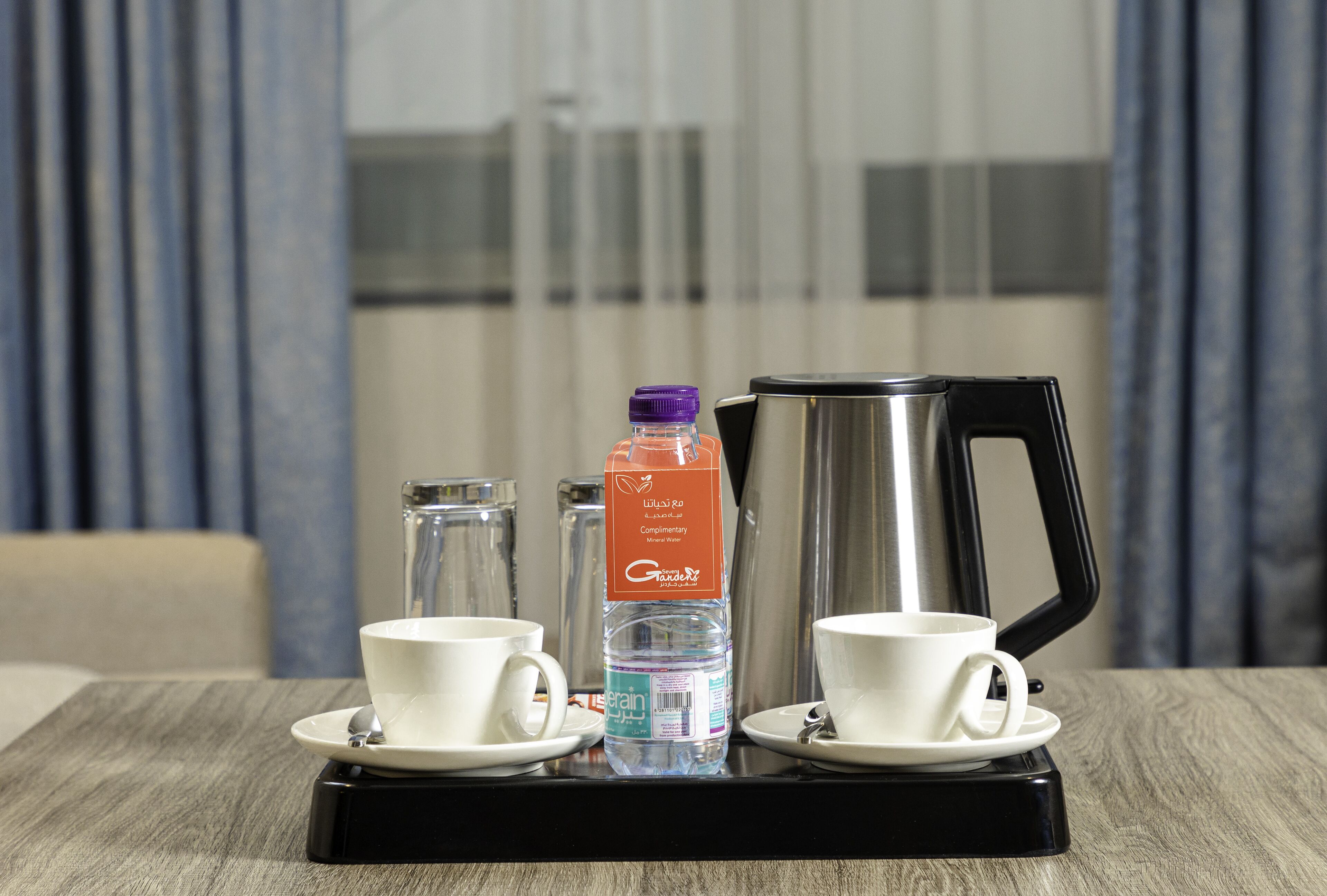 Quarto de hotel com uma mesa posta, com duas chávenas e pires brancos, uma garrafa de água e uma chaleira elétrica de prata.