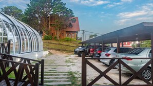 Estrutura de estufa com teto de vidro, carros estacionados e um edifício com telhado vermelho ao fundo.