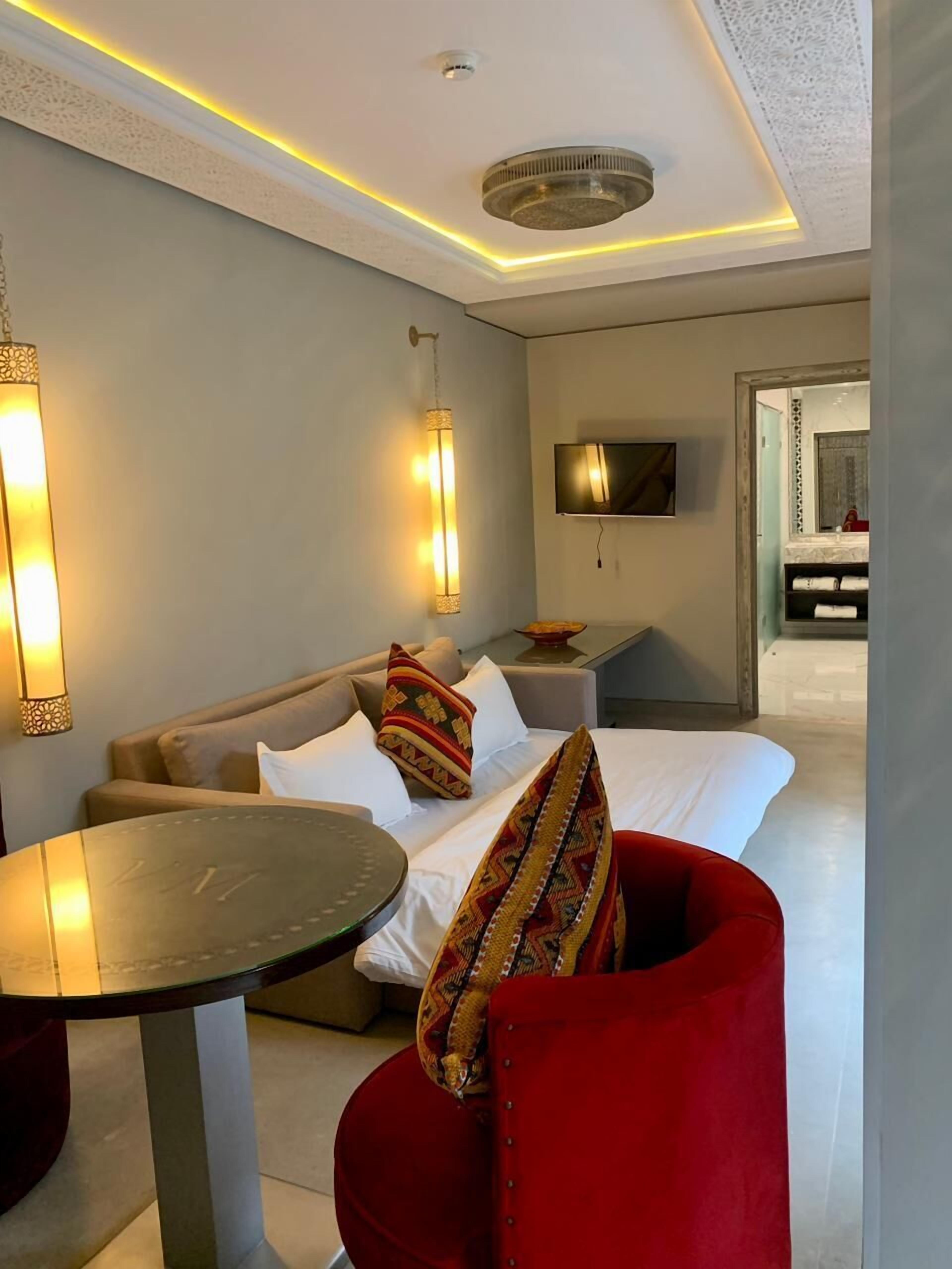 Quarto de hotel moderno com cama, uma cadeira vermelha, uma mesinha e uma TV.