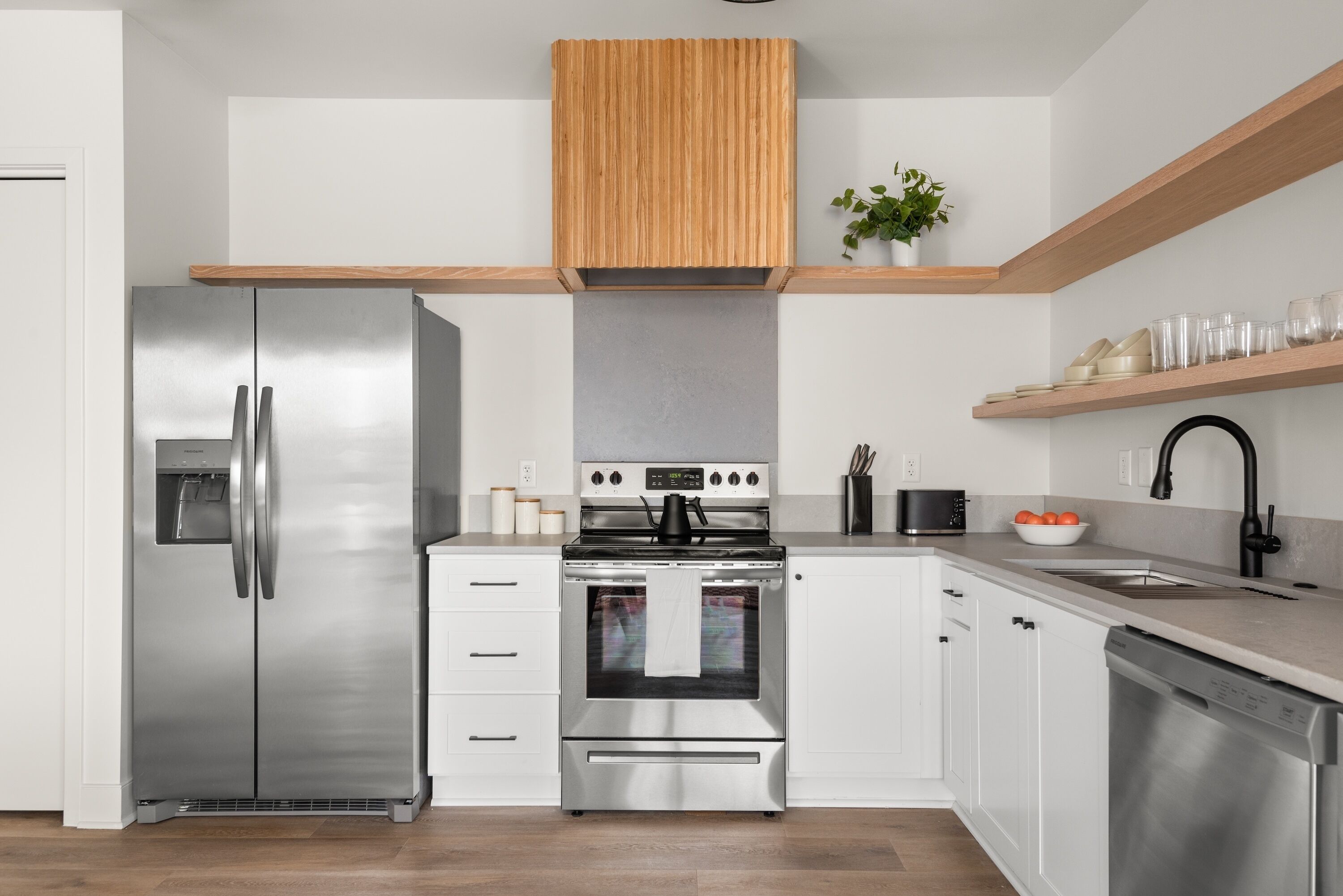 Uma cozinha moderna com geladeira, forno e lava-louças de aço inoxidável, prateleiras de madeira e um vaso de planta.