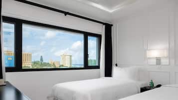 Quarto de hotel com duas camas, uma escrivaninha, uma luminária e vista para edifícios e árvores.