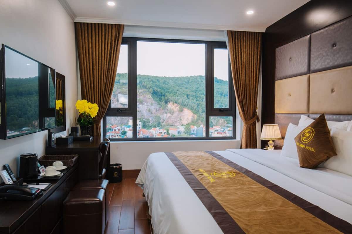 Quarto de hotel com uma cama grande, uma escrivaninha com telefone e chávenas, um candeeiro e vista para uma paisagem montanhosa pela janela.