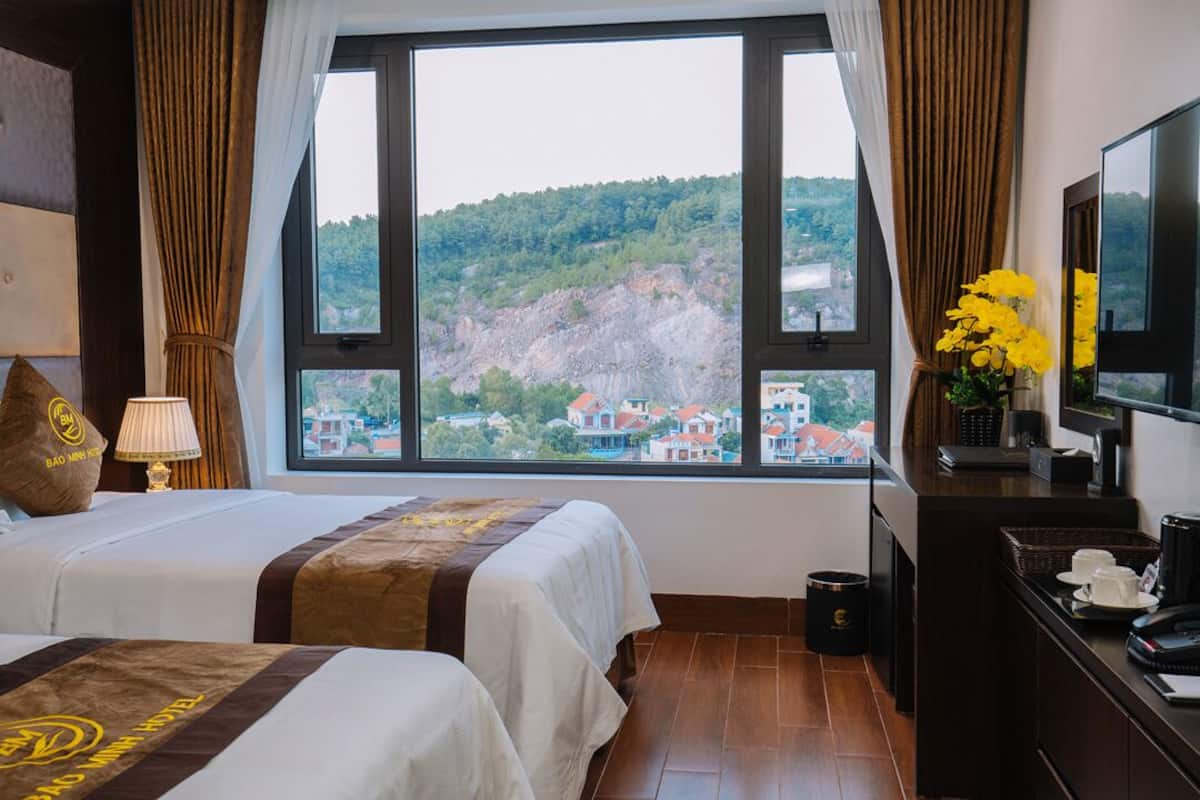 Quarto de hotel com duas camas, uma janela grande com vista para uma colina, uma escrivaninha com televisão e um vaso com flores amarelas.
