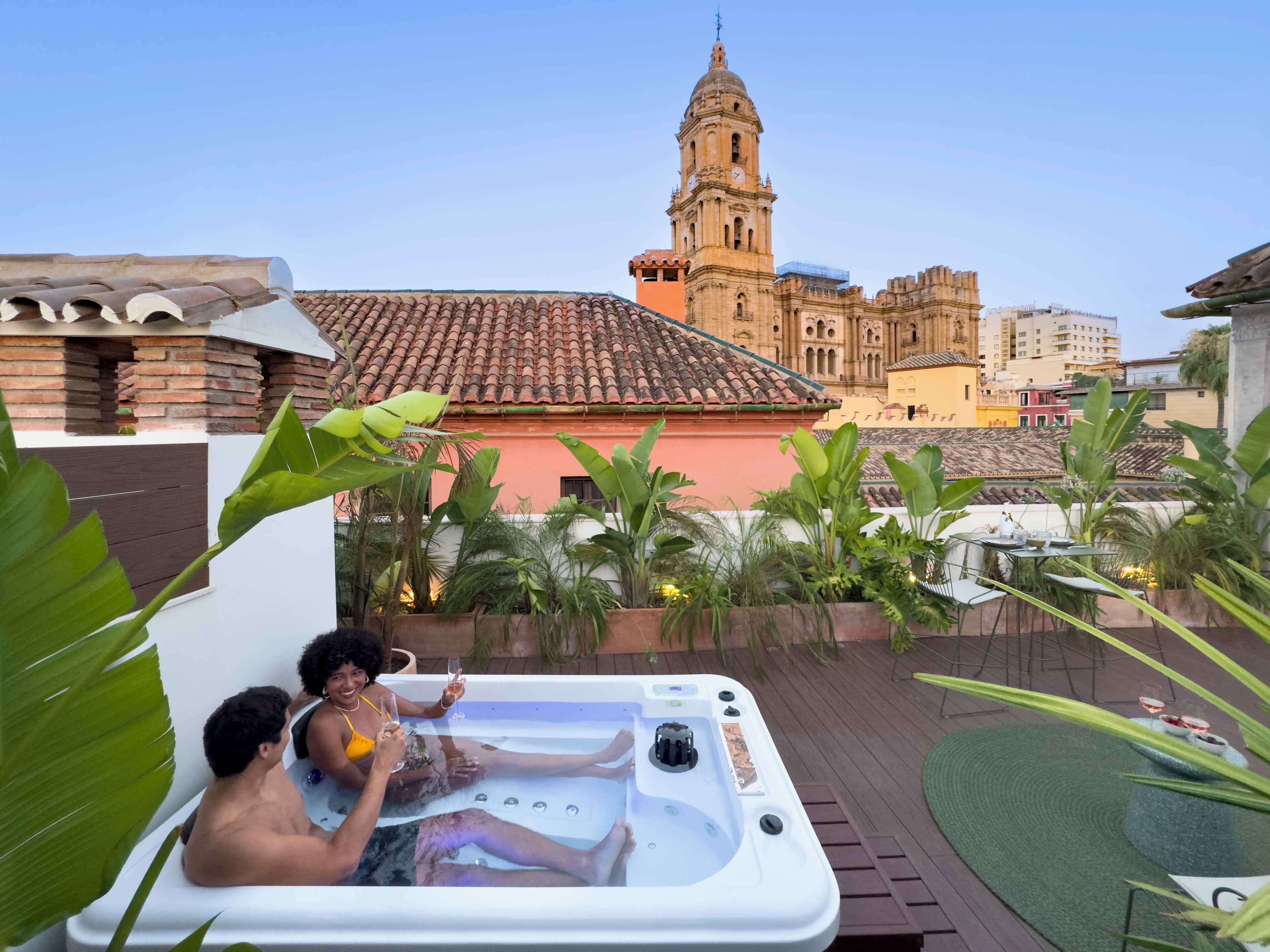 Uma banheira de hidromassagem com duas pessoas, rodeadas por plantas tropicais, com um edifício histórico e uma torre do relógio ao fundo.