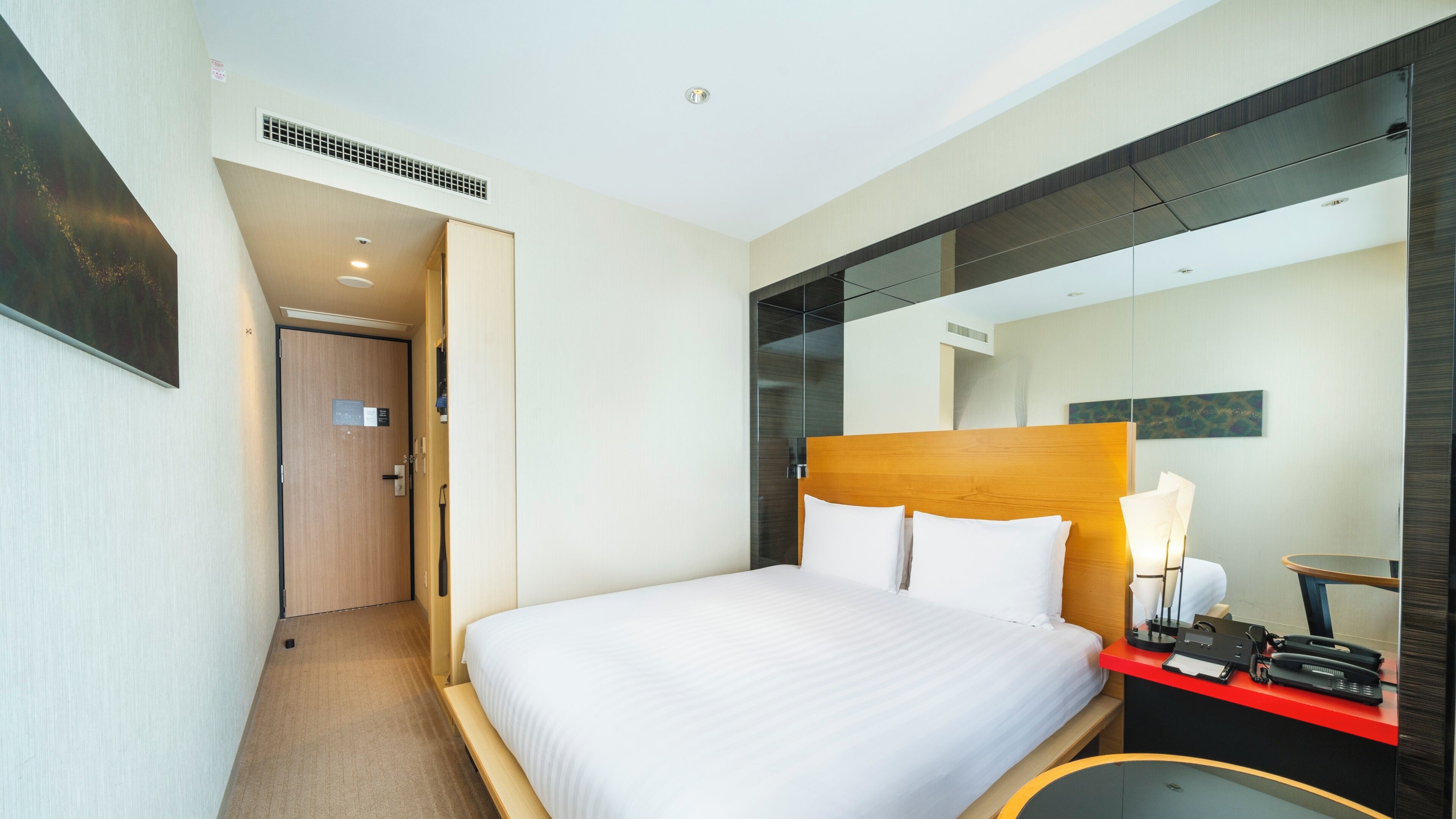 Quarto de hotel com cama, mesa de cabeceira com abajur, espelho e porta para outro cômodo.