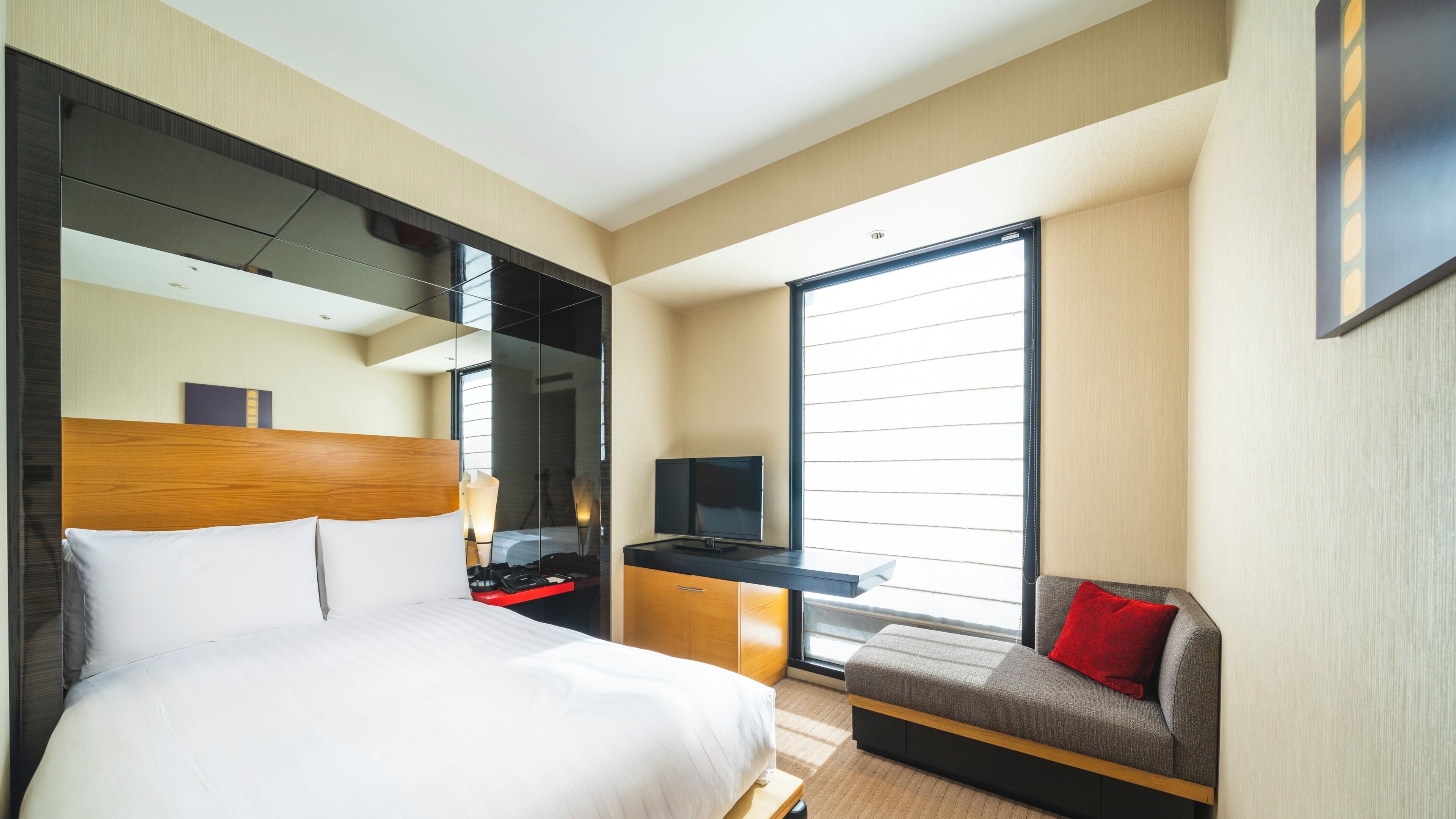 Quarto de hotel com uma cama, uma mesa de trabalho com televisão, um sofá com almofada vermelha e uma janela grande.