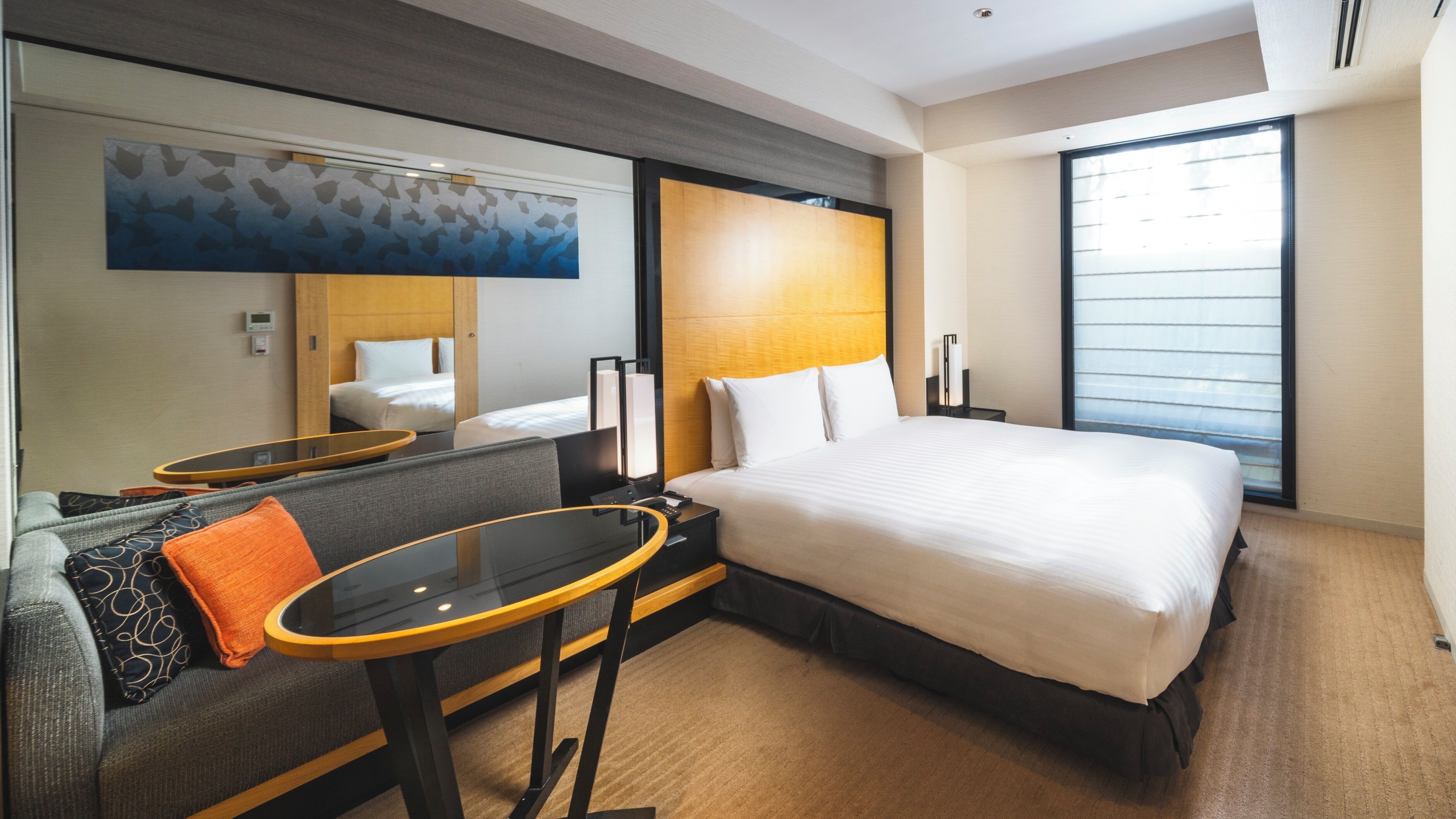 Quarto de hotel com cama, sofá, mesa pequena, espelho e janela grande.