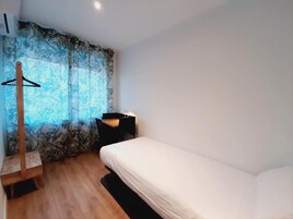 Quarto pequeno e arrumado com uma cama de solteiro, um cabide de madeira, uma escrivaninha com cadeira e uma janela com cortinas estampadas.