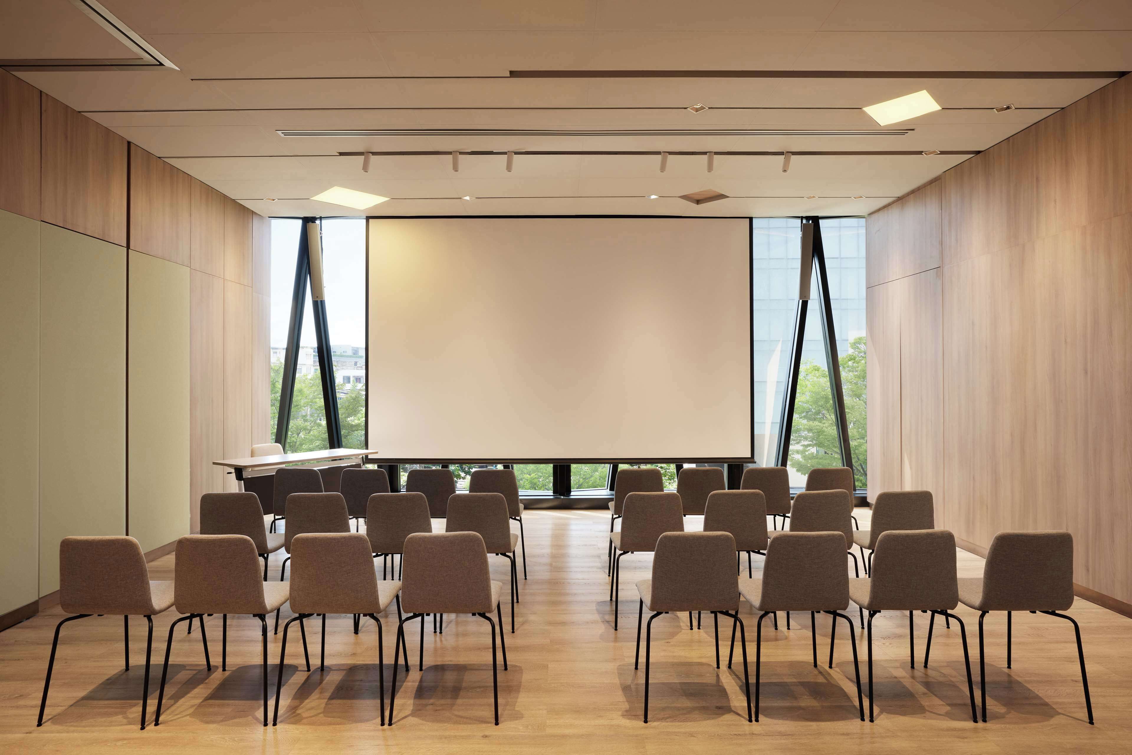 Sala de conferências com uma tela grande, fileiras de cadeiras e piso de madeira.