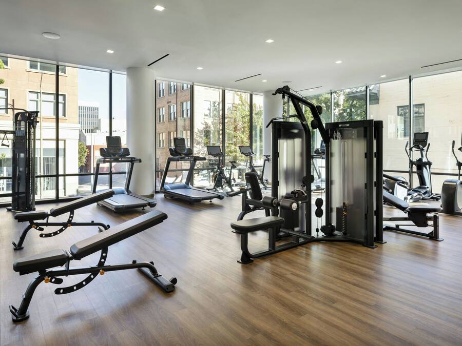 Academia moderna com diversos equipamentos de exercício, janelas amplas e piso de madeira.