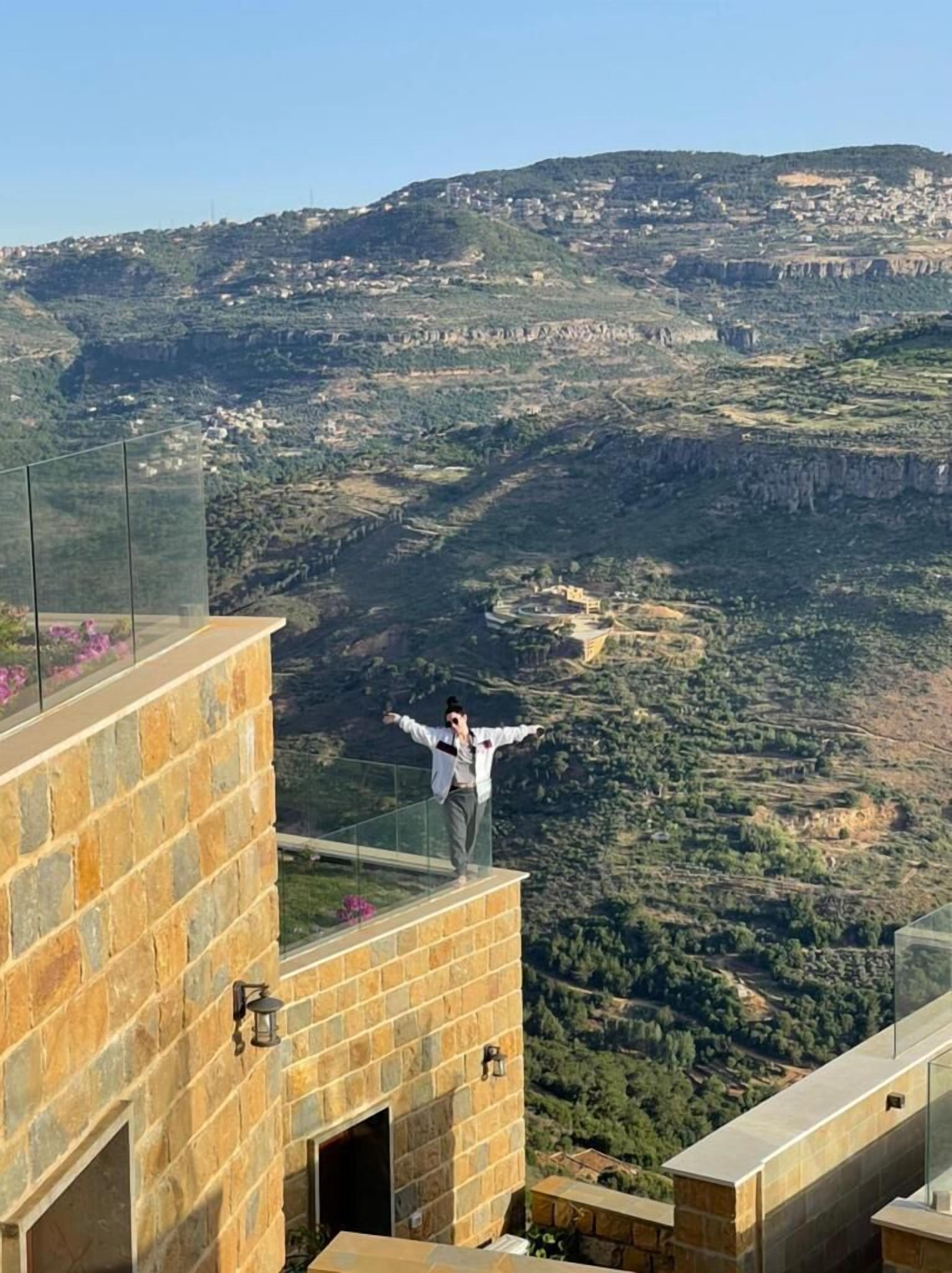 Uma pessoa está em uma varanda, com vista para um vale e edifícios ao longe.
