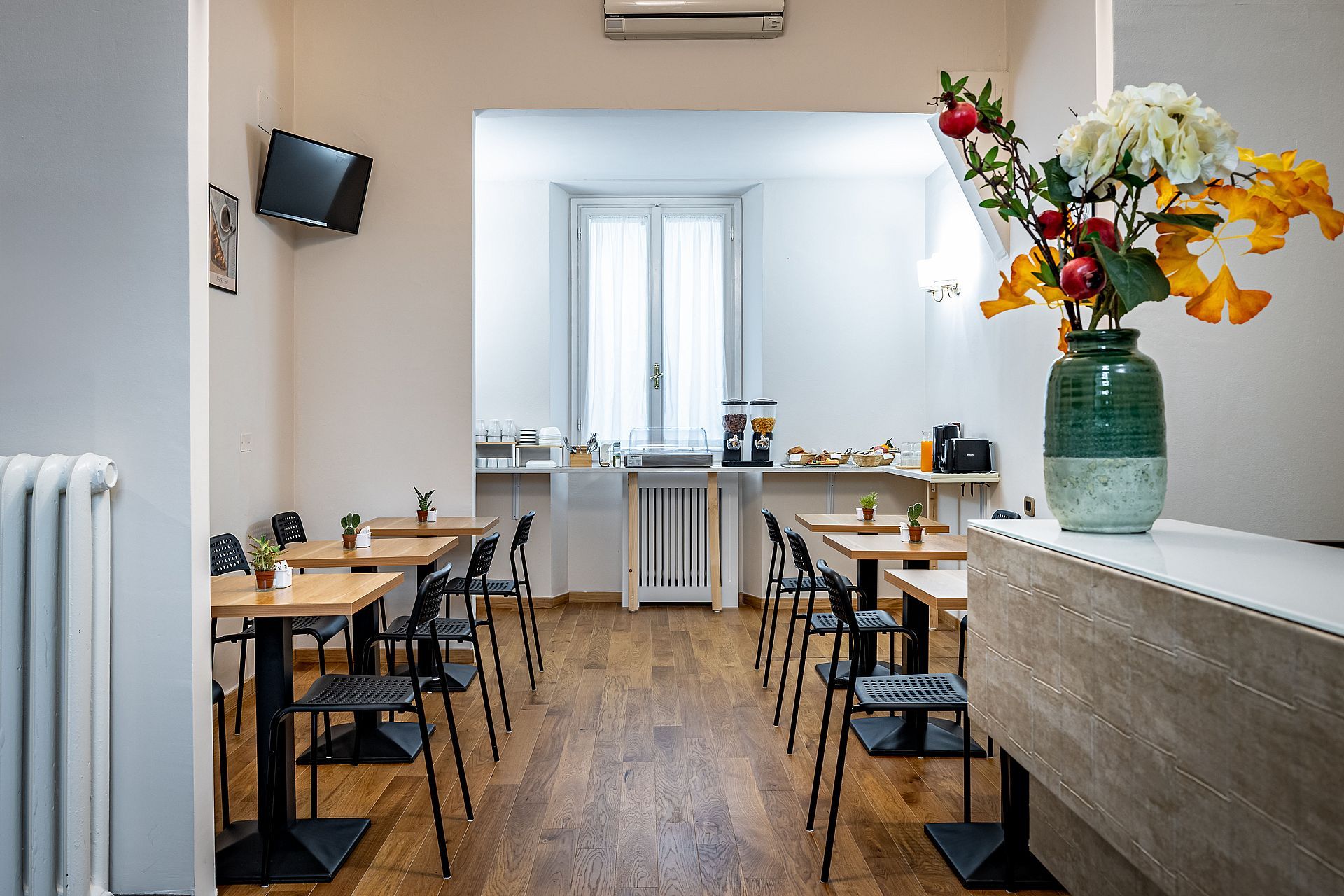 Uma sala de jantar moderna com mesas de madeira e cadeiras pretas, cozinha ao fundo, e um vaso com flores sobre o balcão.