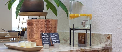 Uma bancada de cozinha com uma planta em vaso, uma panela de cobre, um pote de vidro e um menu de lousa listando bebidas.
