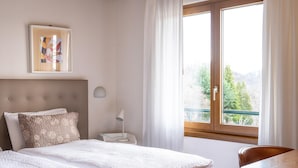 Quarto com cama, mesa de cabeceira, abajur, quadro na parede e janela com vista para árvores.