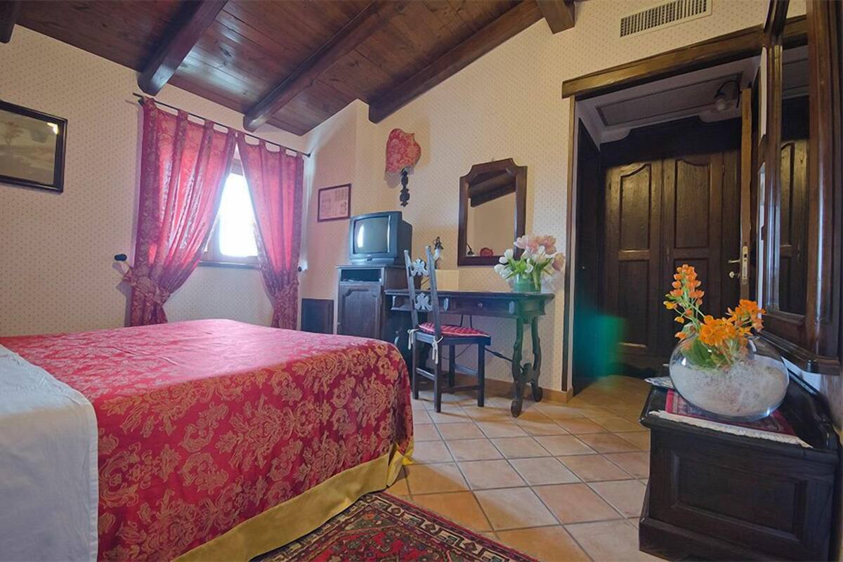 Quarto com cama, escrivaninha, cadeira, televisão, espelho e um vaso com flores.